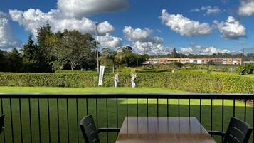 Um campo de golfe com uma mesa e cadeiras em primeiro plano, uma placa com a inscrição 'OXFORD' ao fundo, e um edifício com várias janelas.