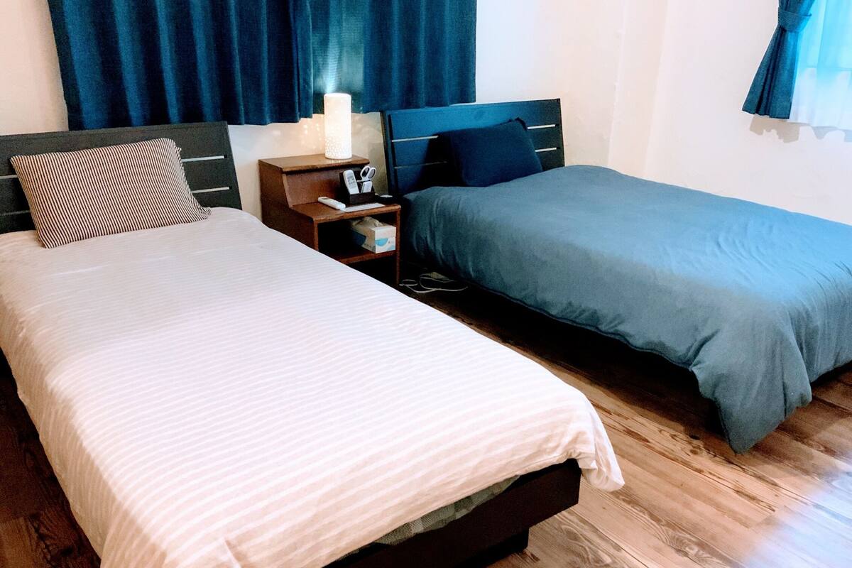 Cama de casal com colcha branca e mesa de cabeceira com abajur, tesoura e controle remoto.