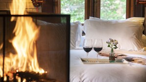 Quarto com lareira, dois copos de vinho e um vaso de flores na cama.