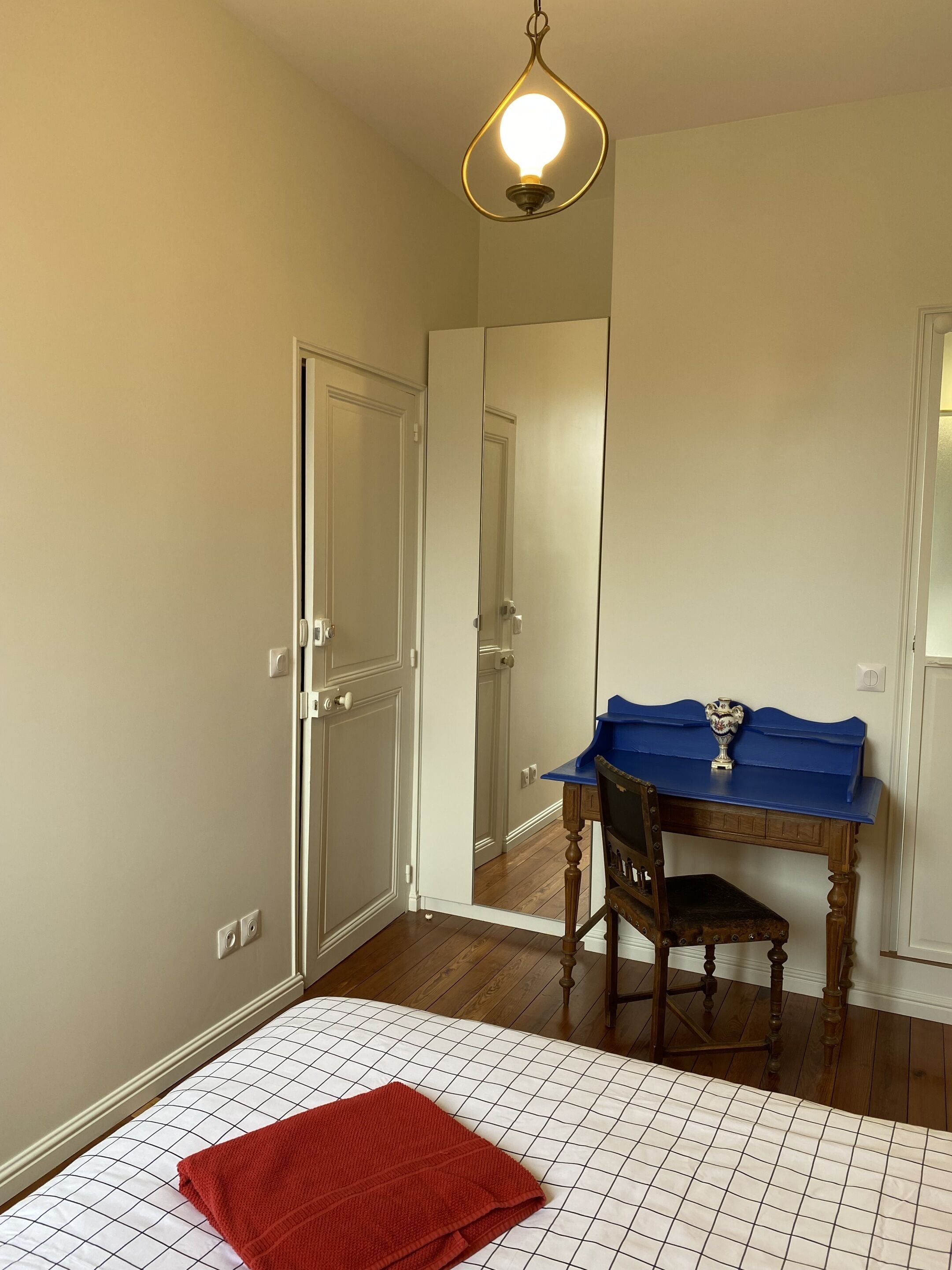 Quarto com cama, escrivaninha e cadeira. Uma porta dá acesso a outro cômodo.