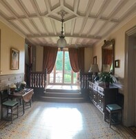Um corredor com ladrilhos estampados, um banco de madeira, uma mesa com um vaso e uma porta que dá para um quarto com uma janela grande e cortinas.