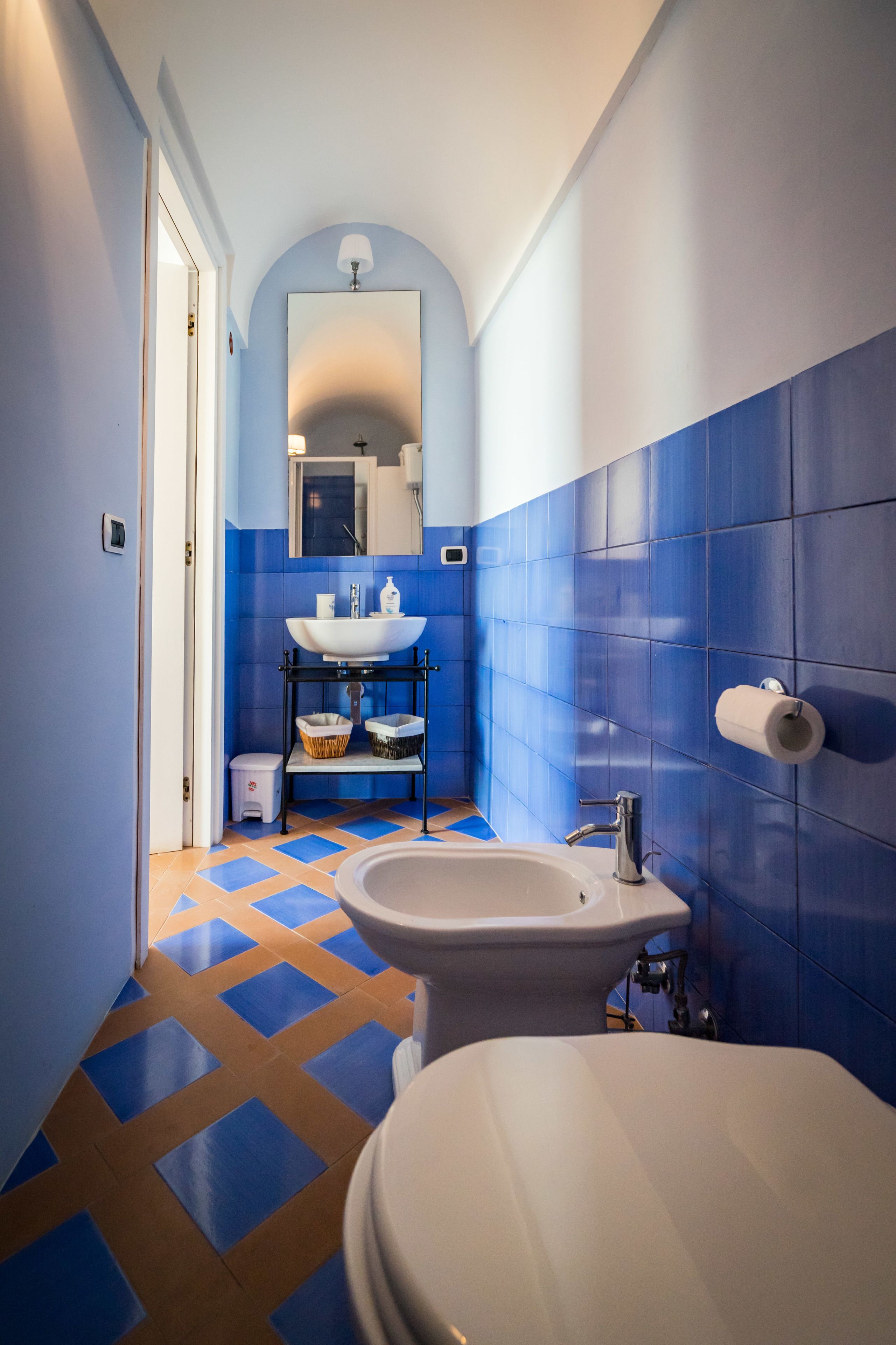 Casa de banho com lavatório branco, sanita e espelho. O chão tem um padrão de azulejos azul e laranja.