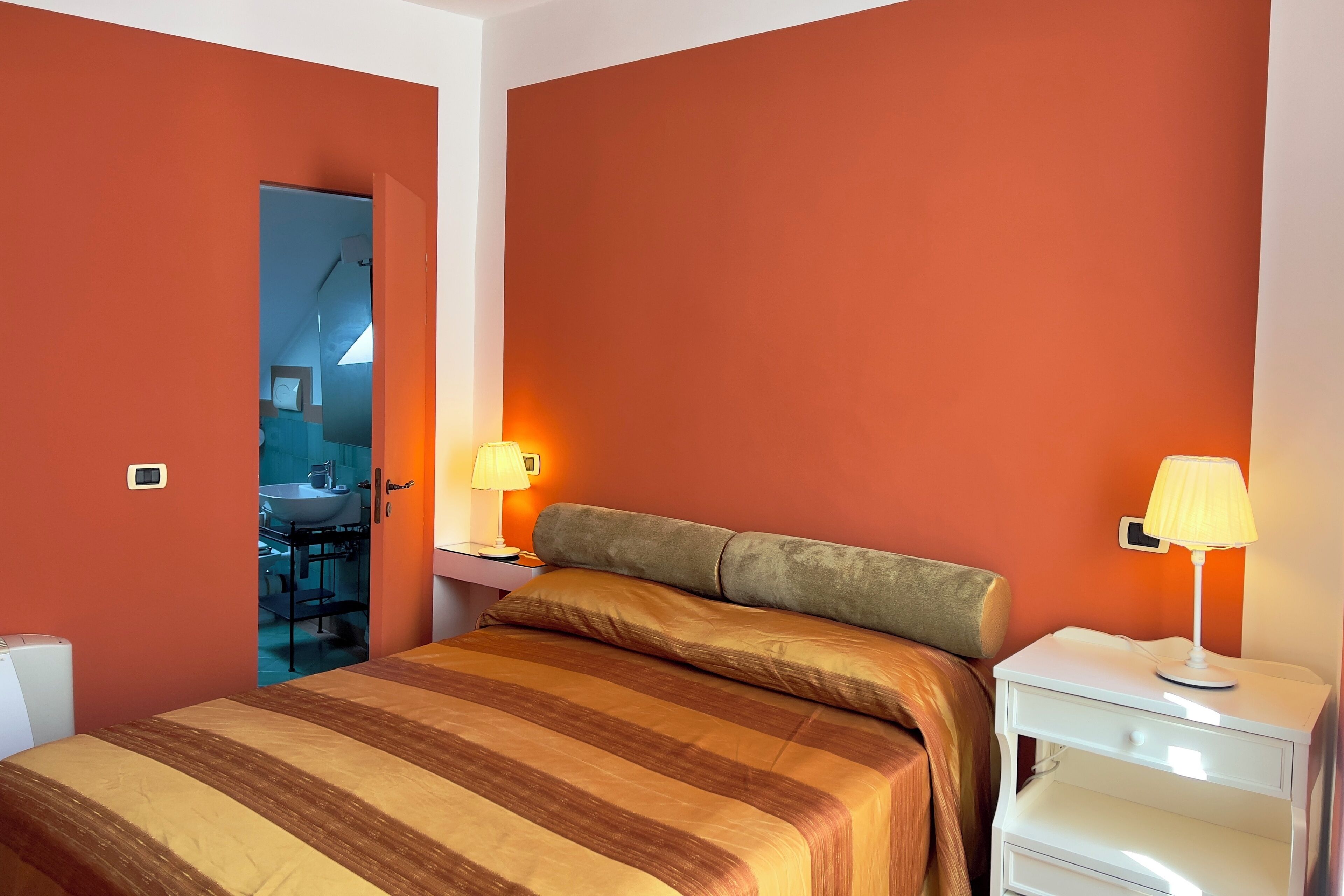 Quarto com uma cama, duas mesas de cabeceira com luminárias, uma porta para um banheiro e um criado-mudo branco.