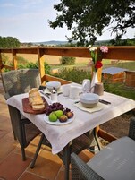 Disposição para refeições ao ar livre com cadeiras de vime, mesa posta com pão, frutas e um vaso de flores.
