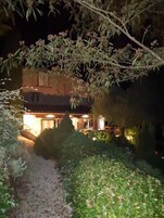 Vista noturna de uma casa com exterior bem iluminado, cercada por vegetação densa e um caminho de cascalho que leva à entrada.