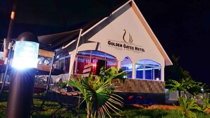 Exterior do Golden Gates Hotel à noite, com uma placa de destaque com o logotipo de um cisne, uma área externa coberta com toldo e uma entrada bem iluminada.