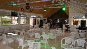 Um espaço amplo e interno com móveis de vime branco, janelas grandes e um palco com equipamentos de iluminação.