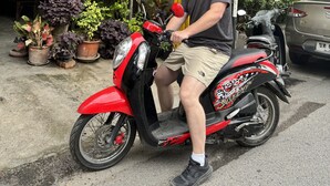Um homem sentado num scooter vermelho estacionado em frente a um prédio com uma placa que diz 'Aunt & Uncle's house Guesthouse'.
