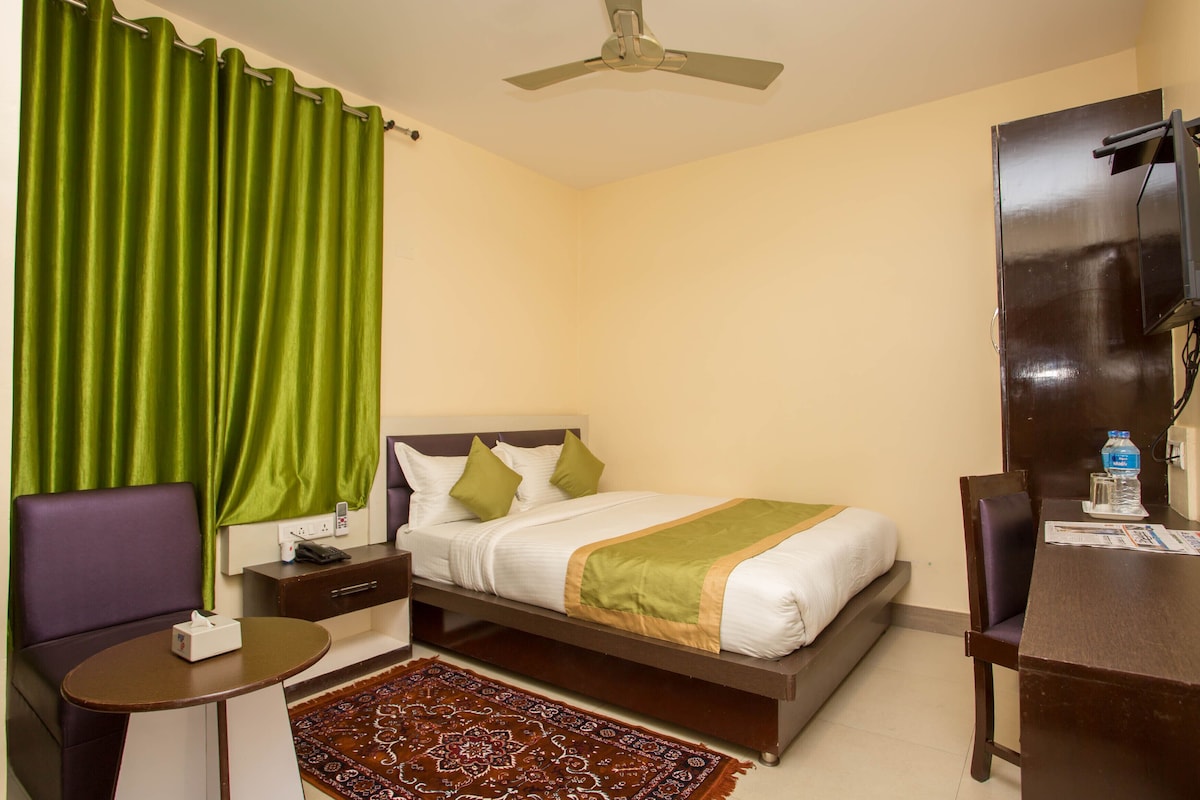 Quarto de hotel com cama, escrivaninha, cadeira, mesa de cabeceira, abajur, ventilador de teto e cortina verde.