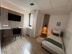 Quarto de hotel com cama, escrivaninha com cadeira, televisão fixada na parede, mesa de cabeceira com abajur e janela com cortinas.