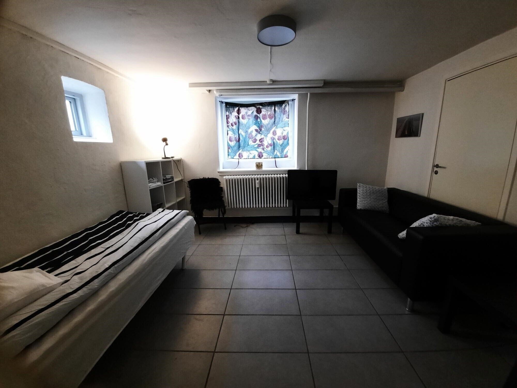 Quarto pequeno e pouco mobiliado, com cama, sofá, radiador e janela com cortinas florais.
