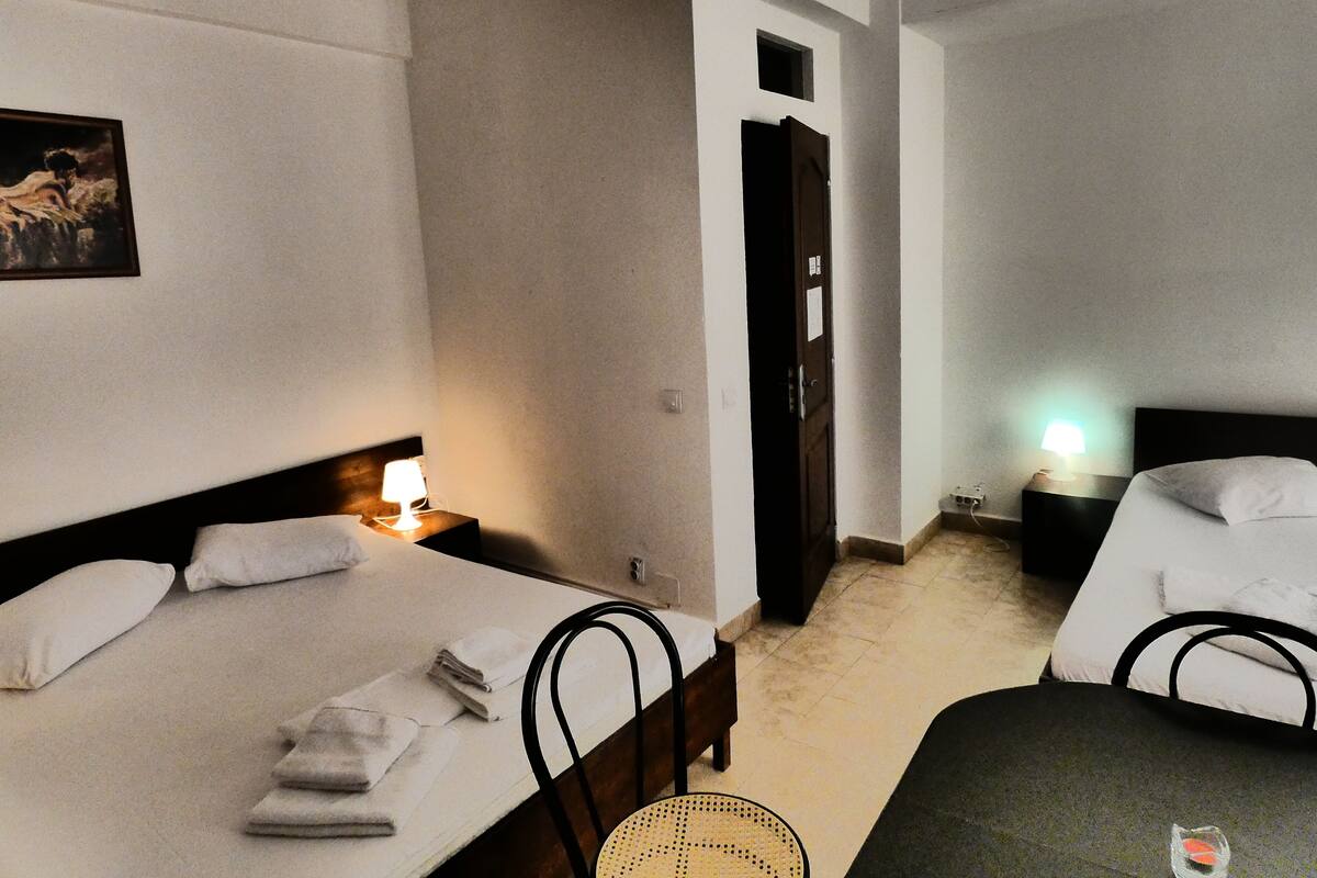 Quarto de hotel com duas camas, cabeceira, mesa de cabeceira com abajur, quadro na parede e uma porta.