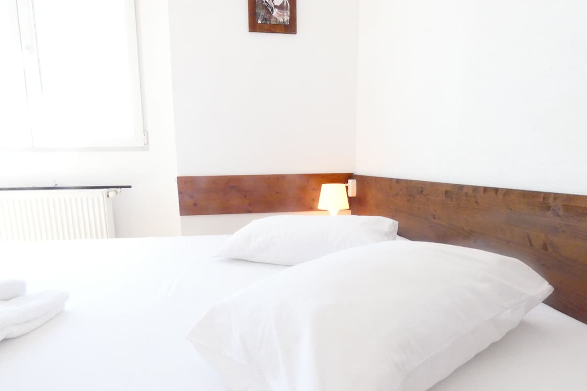Cama bem arrumada com lençóis e almofadas brancos, cabeceira de madeira, abajur e quadro na parede.