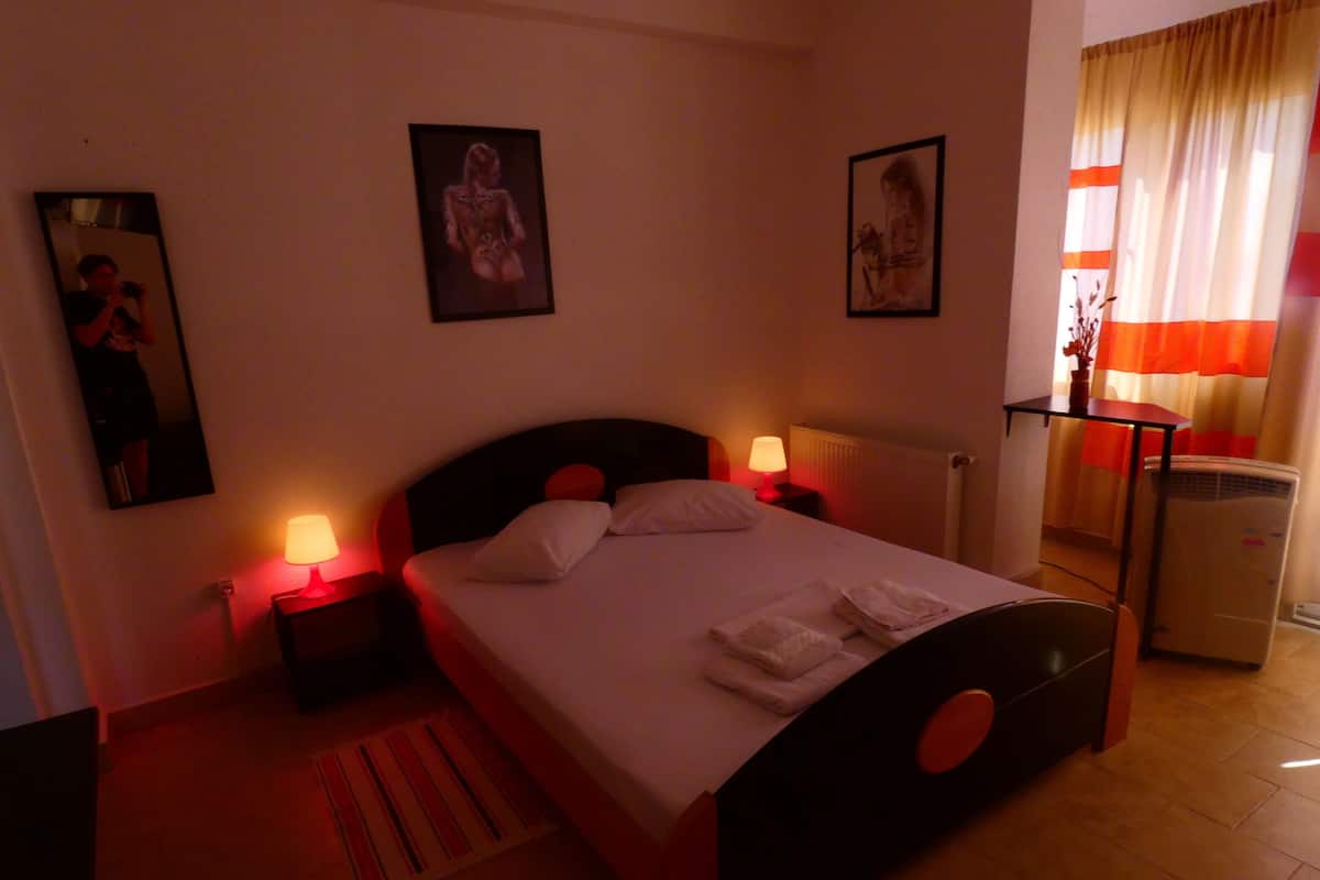 Quarto com cama, duas mesas de cabeceira com luminárias, radiador, janela com cortinas e um cesto de lixo.