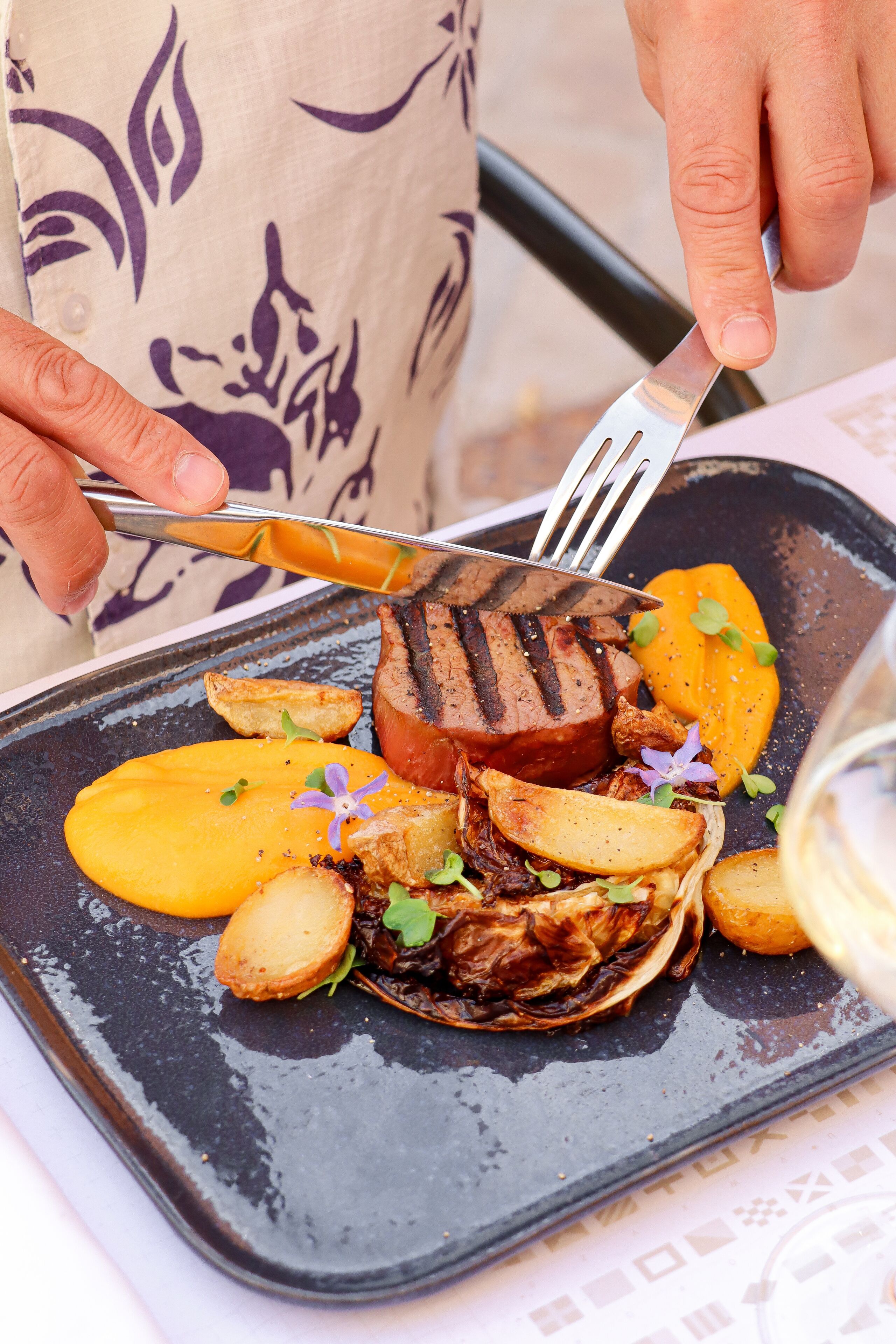 Uma pessoa cortando um bife com garfo e faca em um prato com batatas e um legume amarelo.