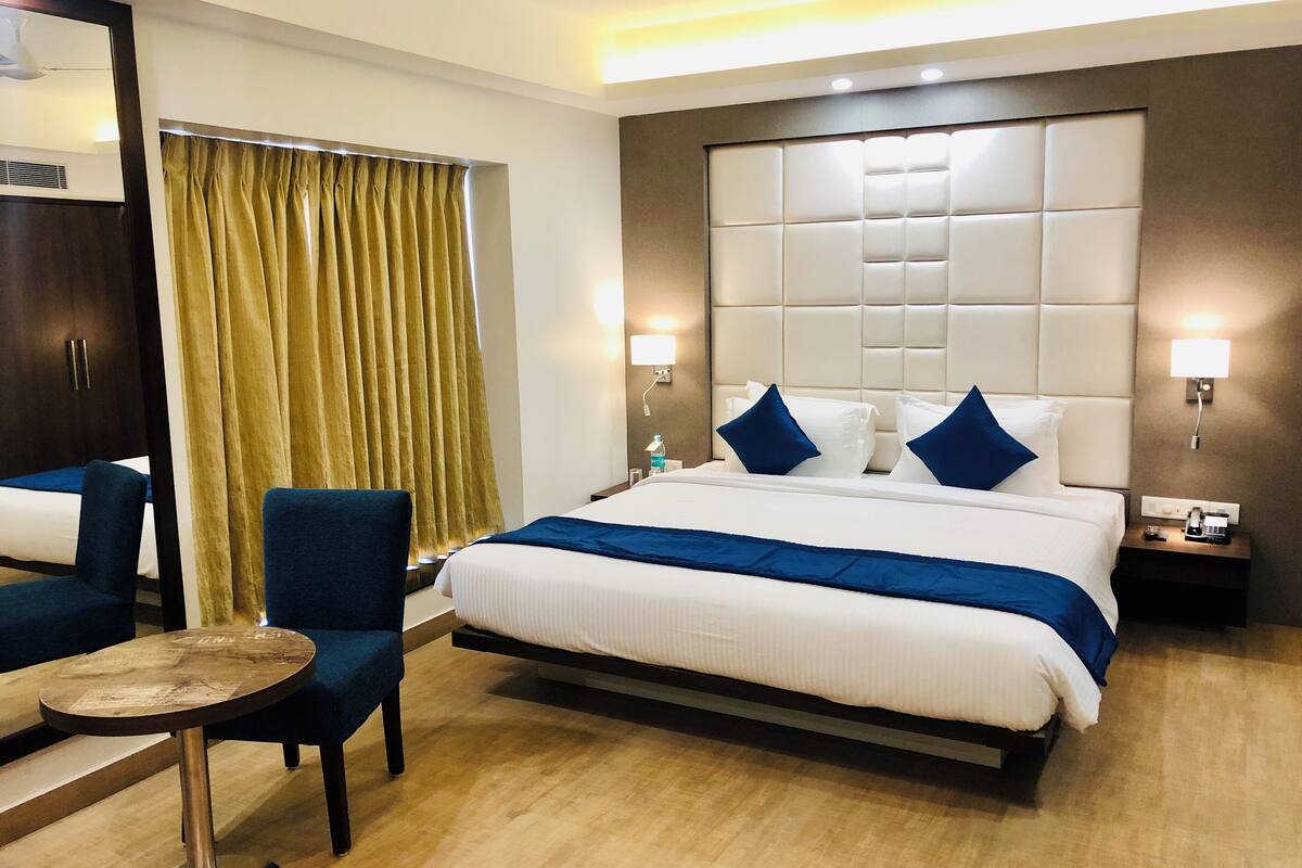 Quarto de hotel com uma cama grande, uma mesa com cadeira, um espelho e uma mesinha de cabeceira com telefone.