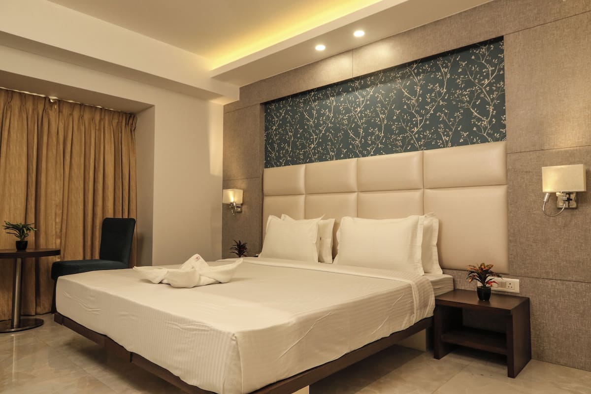 Quarto de hotel moderno com uma cama grande, cabeceira com padrão floral, mesa de cabeceira com luminária e uma poltrona.
