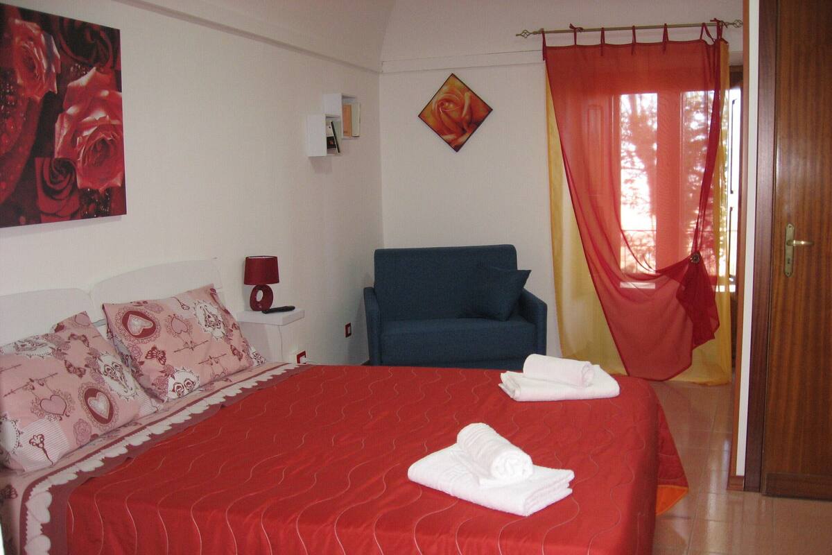 Cama bem arrumada com roupa de cama vermelha e toalhas brancas, uma poltrona azul, uma mesinha de cabeceira com abajur, e um quadro de rosas na parede.