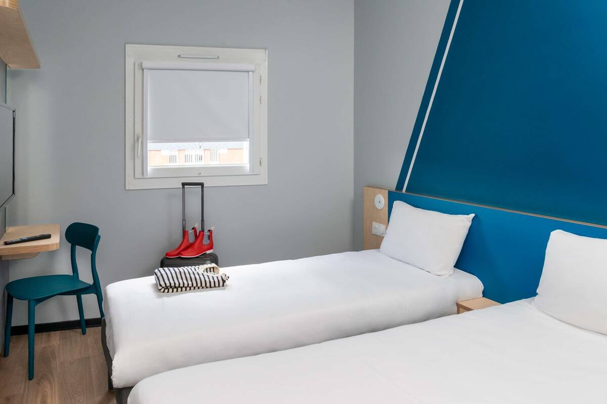 Quarto de hotel com duas camas, uma escrivaninha, uma cadeira, uma mala e uma janela.