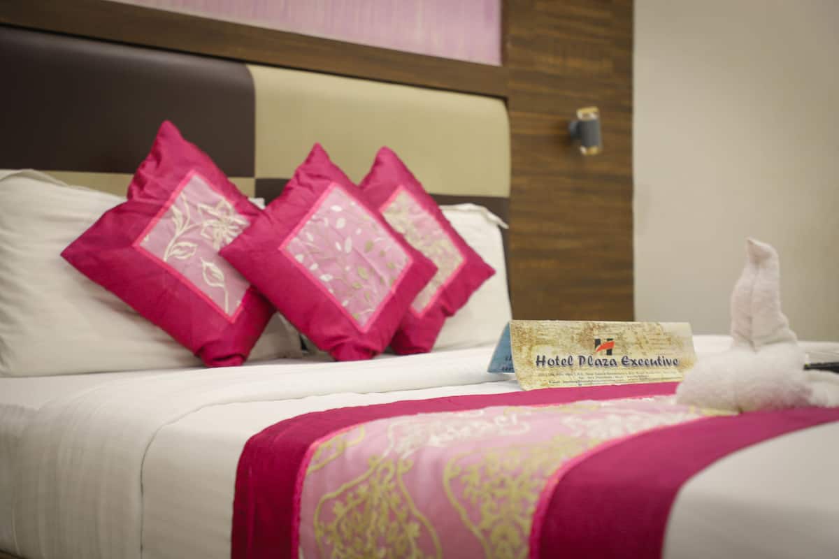 Quarto de hotel com uma cama, almofadas rosa e brancas, uma almofada branca e fofa, e um letreiro com a inscrição "Hotel Plaza Executive".