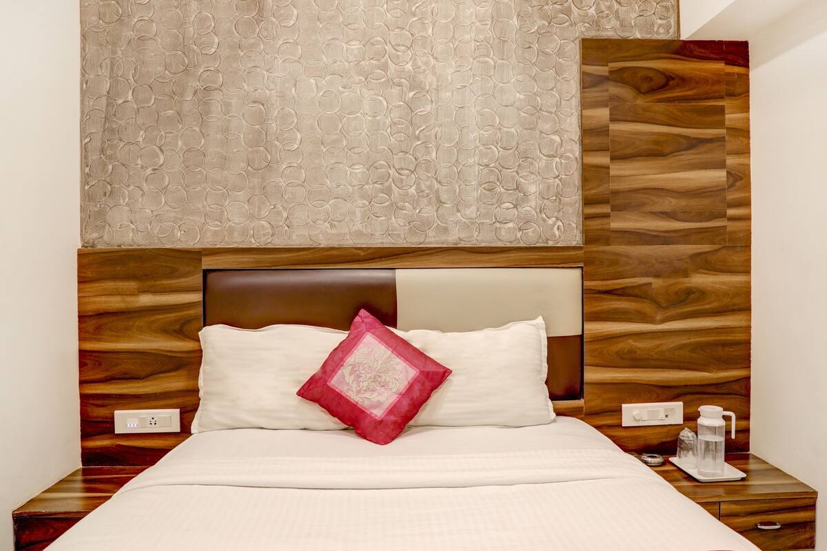 Cama bem arrumada com cabeceira de madeira, travesseiro decorativo vermelho e mesa de cabeceira com garrafa de água e um celular.