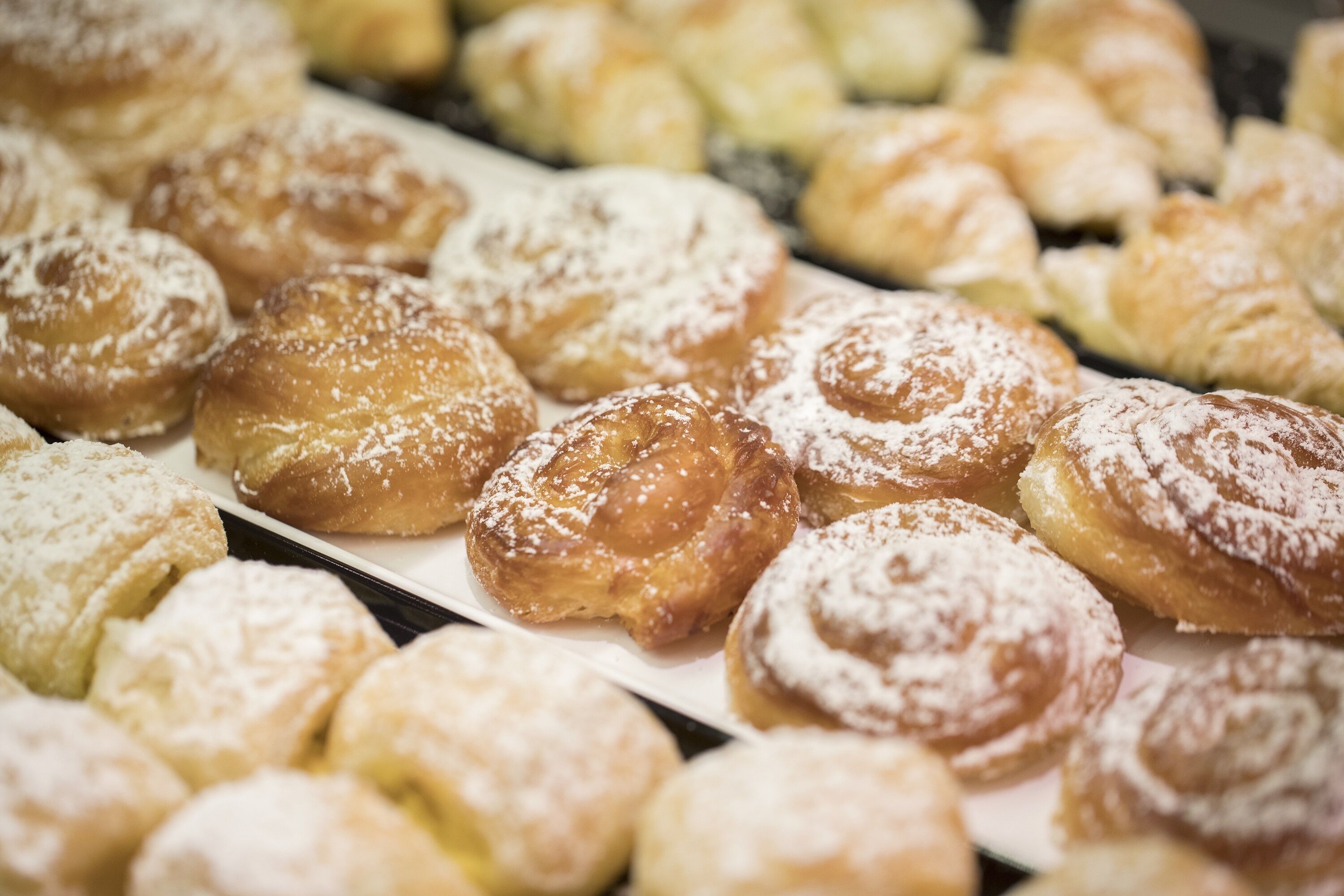 Exposição de diversos doces, incluindo donuts e pastéis polvilhados com açúcar de confeiteiro.