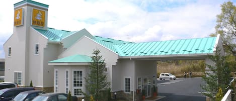 Hotel com telhado verde e torre com sinalização. Carros estacionados em frente e algumas pessoas caminhando.