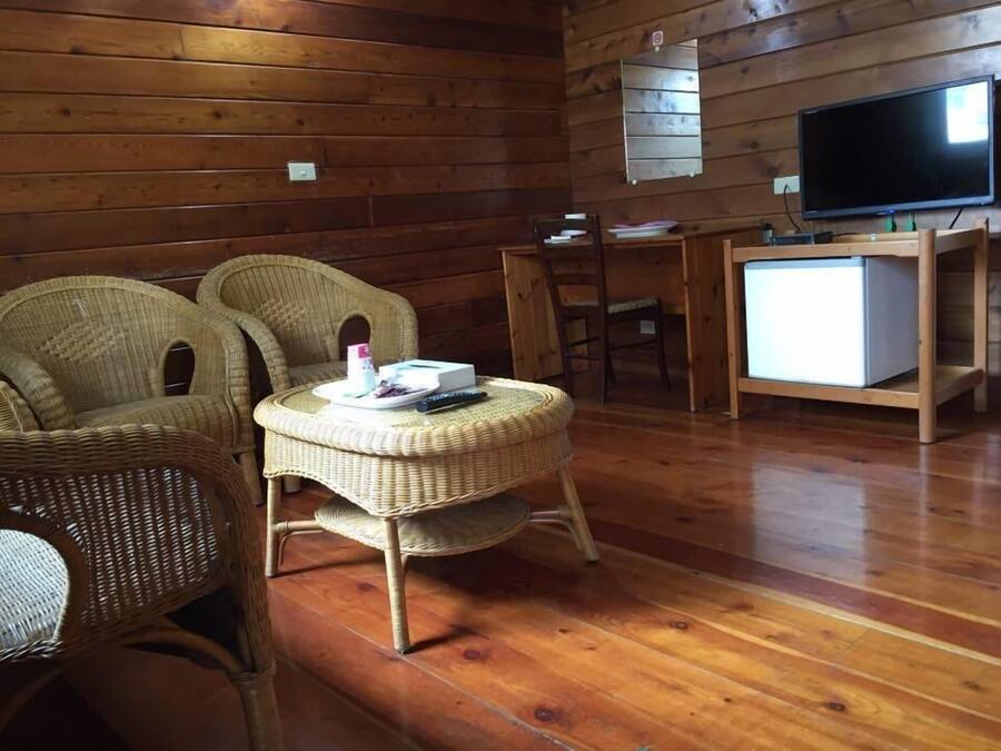 Quarto aconchegante com paredes e pisos de madeira, com duas cadeiras de vime, uma mesa pequena com um controle remoto, uma televisão e um refrigerador.