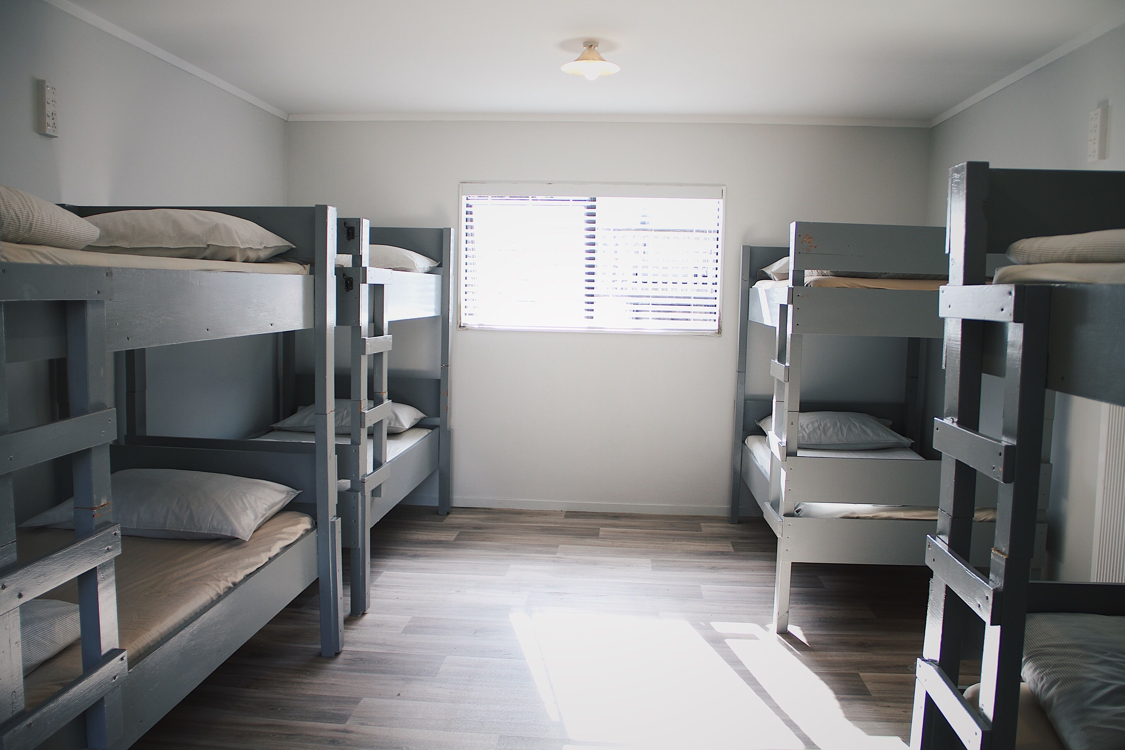Quarto com beliches dispostos em duas fileiras, janela com persianas e luminária no teto.