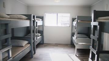 Quarto com beliches dispostos em duas fileiras, janela com persianas e luminária no teto.