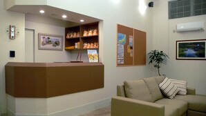 Uma área de recepção com balcão marrom, um quadro emoldurado, um mural de avisos e um vaso de planta.