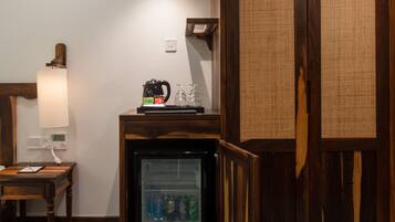 Quarto de hotel compacto com mini-geladeira repleta de bebidas, cafeteira e mesa com luminária.