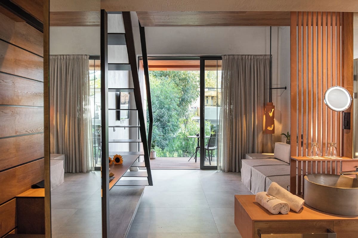 Quarto de hotel moderno com uma grande porta de vidro deslizante que dá para uma varanda, um banco de madeira, um espelho redondo e uma pia espaçosa.