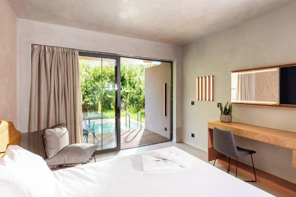 Quarto de hotel moderno com uma cama, escrivaninha, cadeira, televisão e porta de vidro que dá para uma área de piscina.