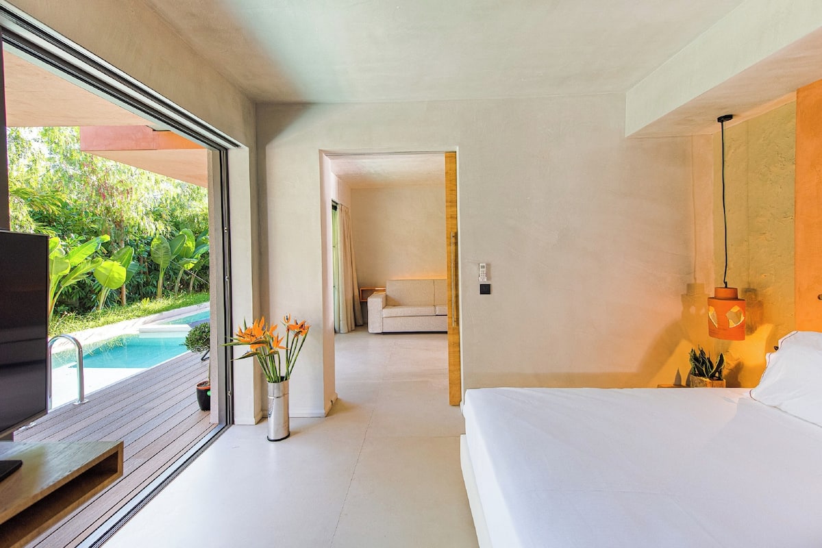 Quarto de hotel moderno com uma cama grande, televisão, mesa pequena e vista para área de piscina externa.