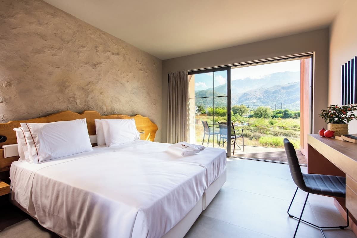 Quarto com uma cama grande, uma escrivaninha e vista para montanhas através de uma porta de correr de vidro.