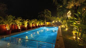 Piscina à noite, com palmeiras ao redor e um edifício ao fundo.