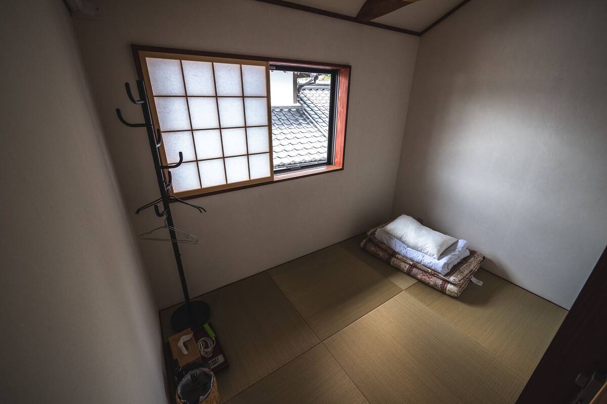 Quarto com piso de tatame, cama futon, janela com tela shoji e uma pequena mesa com um cesto.