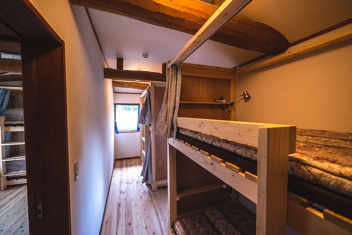 Quarto pequeno com chão de madeira, uma cama beliche, uma janela e uma escrivaninha com abajur.