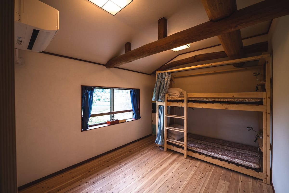 Um quarto com uma cama beliche, uma janela com cortinas, piso de madeira e vigas de madeira aparentes.