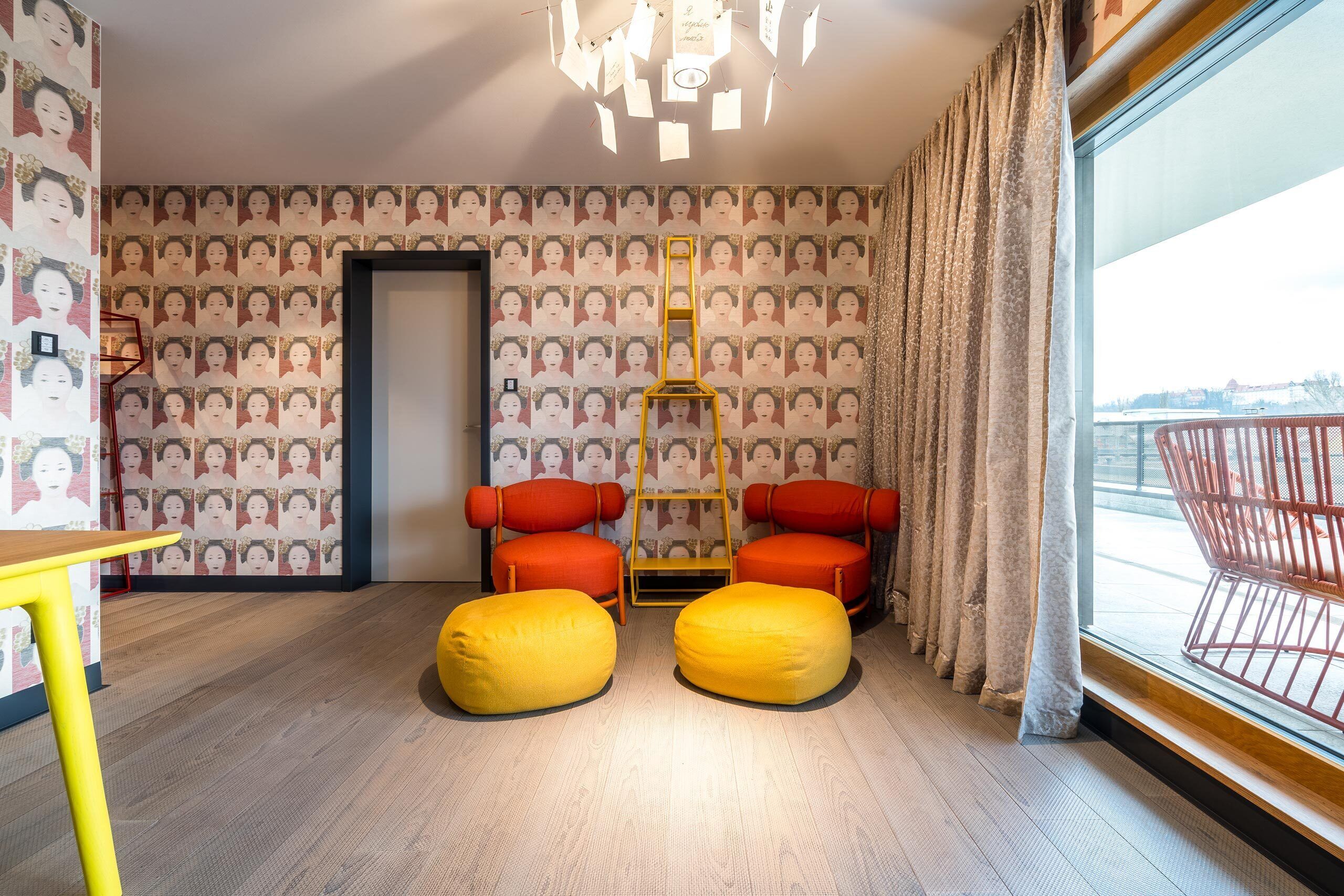 Um quarto com papel de parede estampado, piso de madeira, uma escada amarela, duas cadeiras laranjas e um banquinho amarelo.