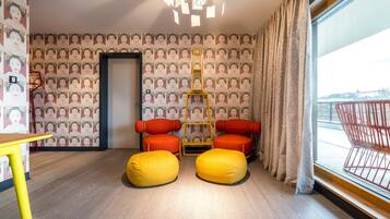 Um quarto com papel de parede estampado, piso de madeira, uma escada amarela, duas cadeiras laranjas e um banquinho amarelo.