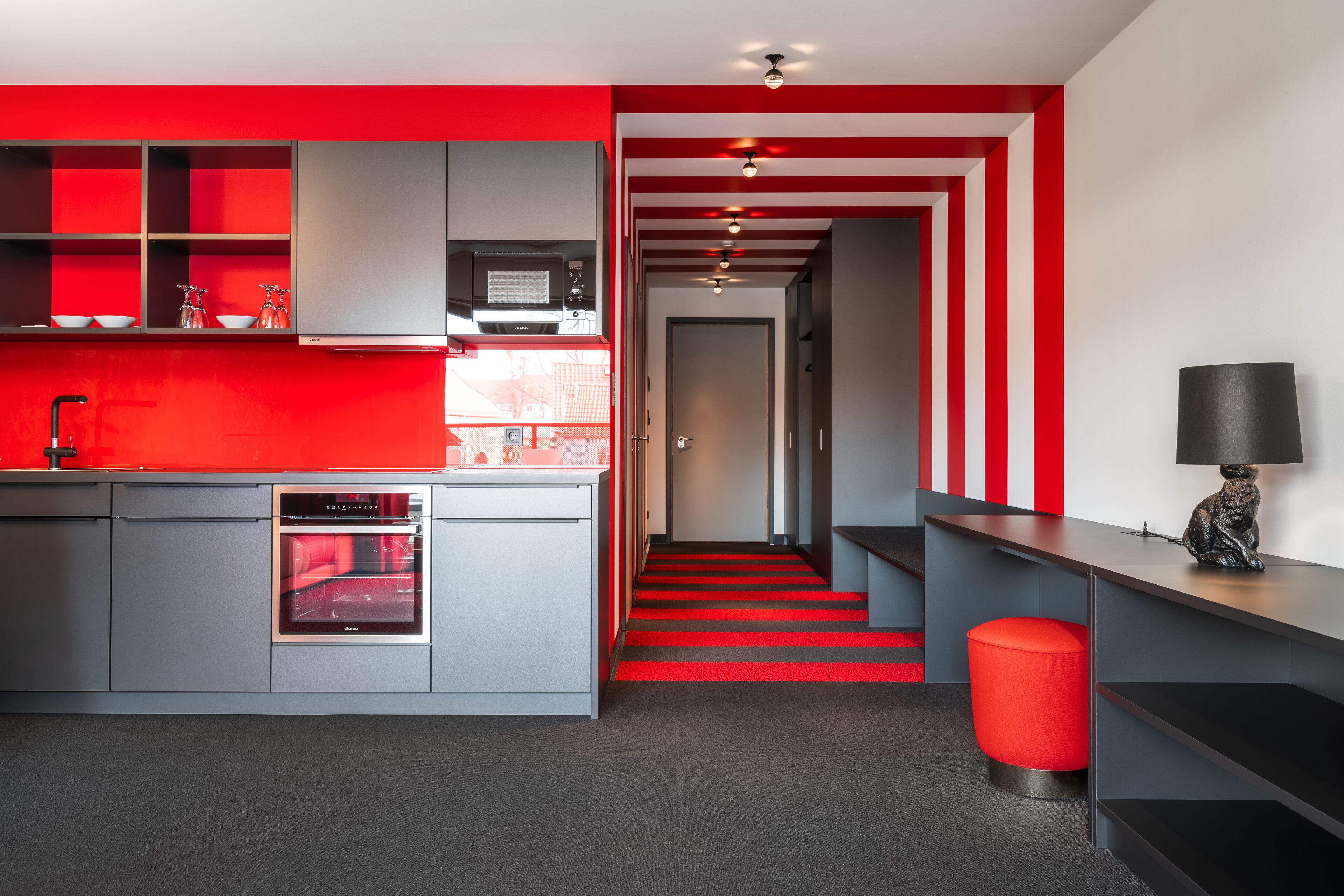 Cozinha moderna com armários cinza, bancada vermelha e escada listrada de vermelho que leva a um corredor.