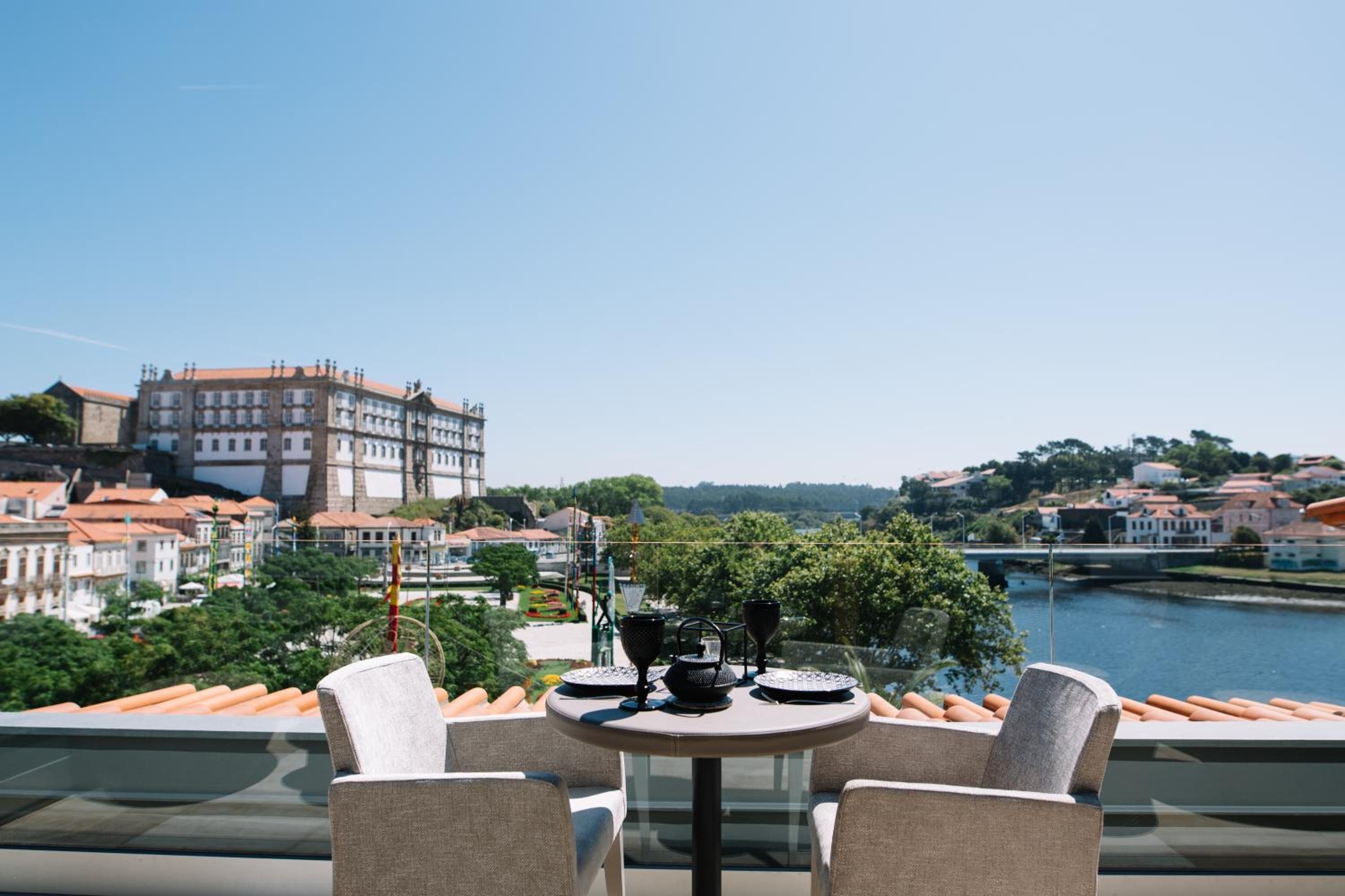 Mesa com cadeiras numa varanda com vista para um rio, com edifícios ao fundo.