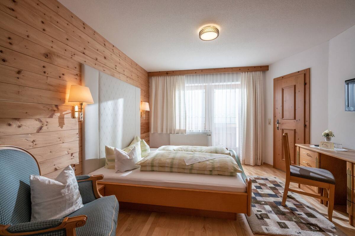 Um quarto aconchegante com cabeceira de madeira, cama com roupa de cama xadrez, escrivaninha com cadeira e janela com cortinas leves.
