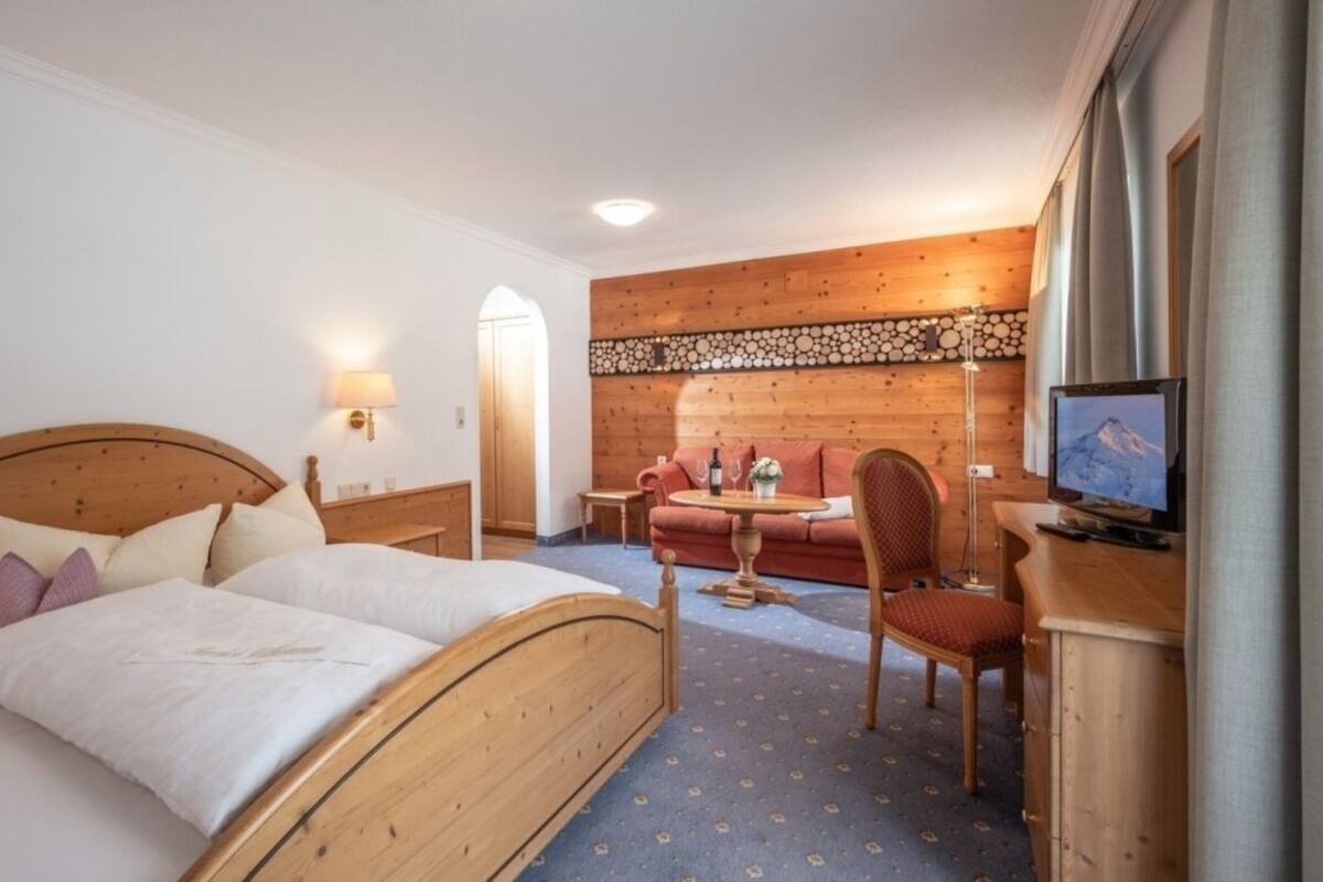 Quarto de hotel com cama de madeira, um sofá, uma televisão e uma pequena mesa com abajur.