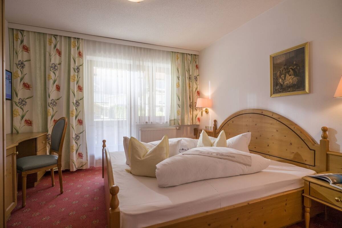 Quarto de hotel com cama de madeira, cortinas florais, escrivaninha e luminária.