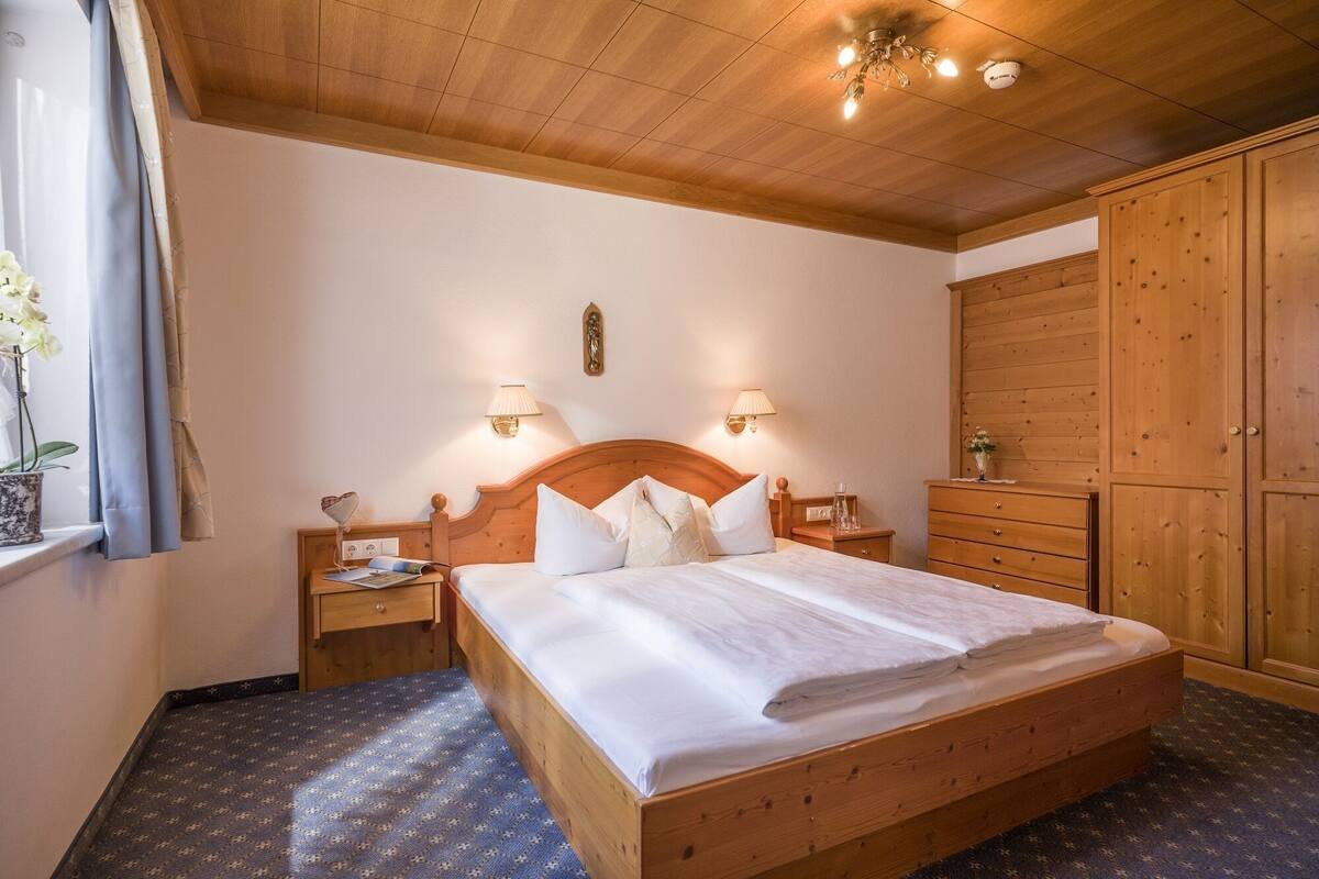 Uma cama com estrutura de madeira, roupa de cama branca, duas luminárias de cabeceira, uma mesa de cabeceira e um guarda-roupa de madeira num quarto com teto inclinado e carpete estampado.