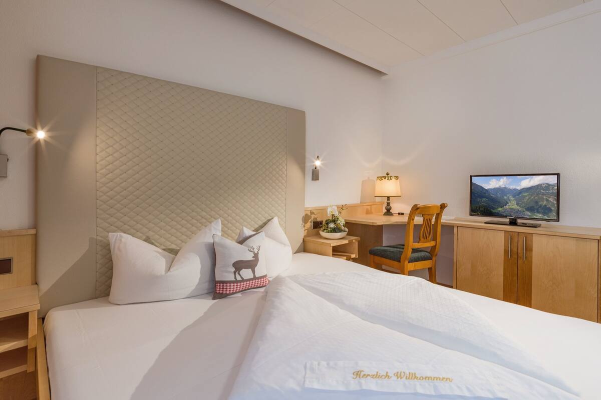 Quarto de hotel com cama, escrivaninha, cadeira, televisão e luminária na parede.