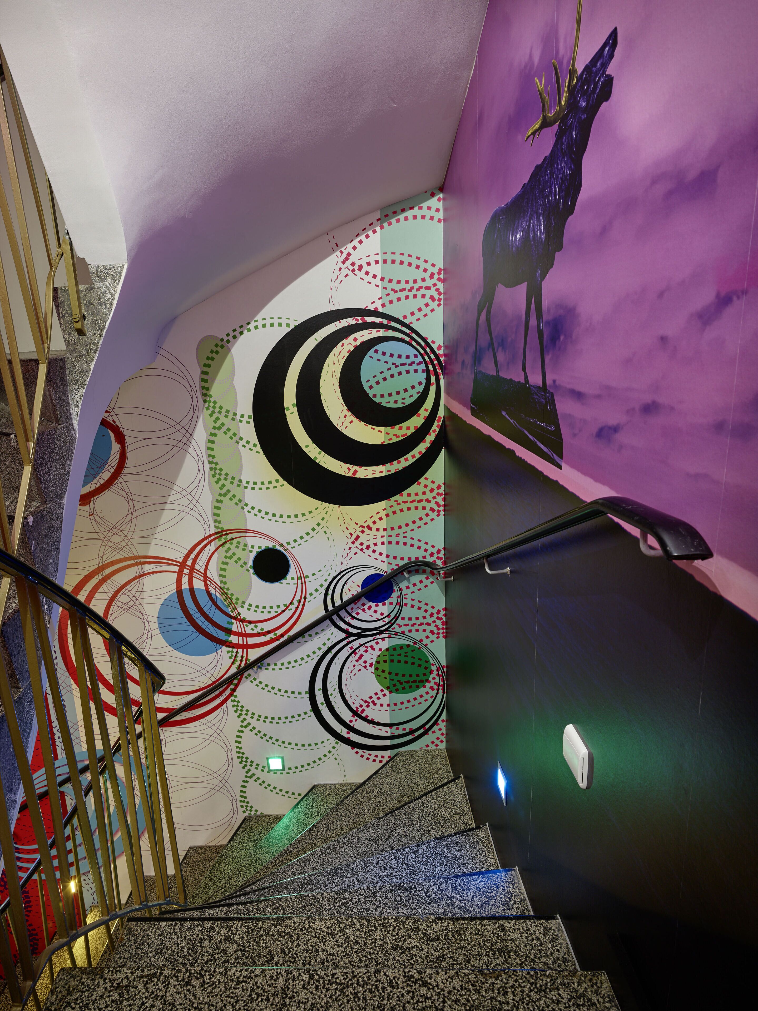 Uma escadaria com um mural abstrato e colorido na parede e uma pintura de um veado à direita.